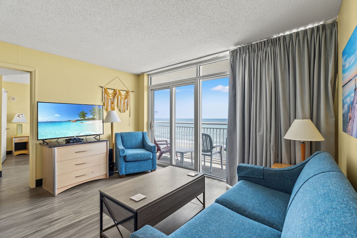 Quarto de hotel com varanda, televisão, sofá e secretária.