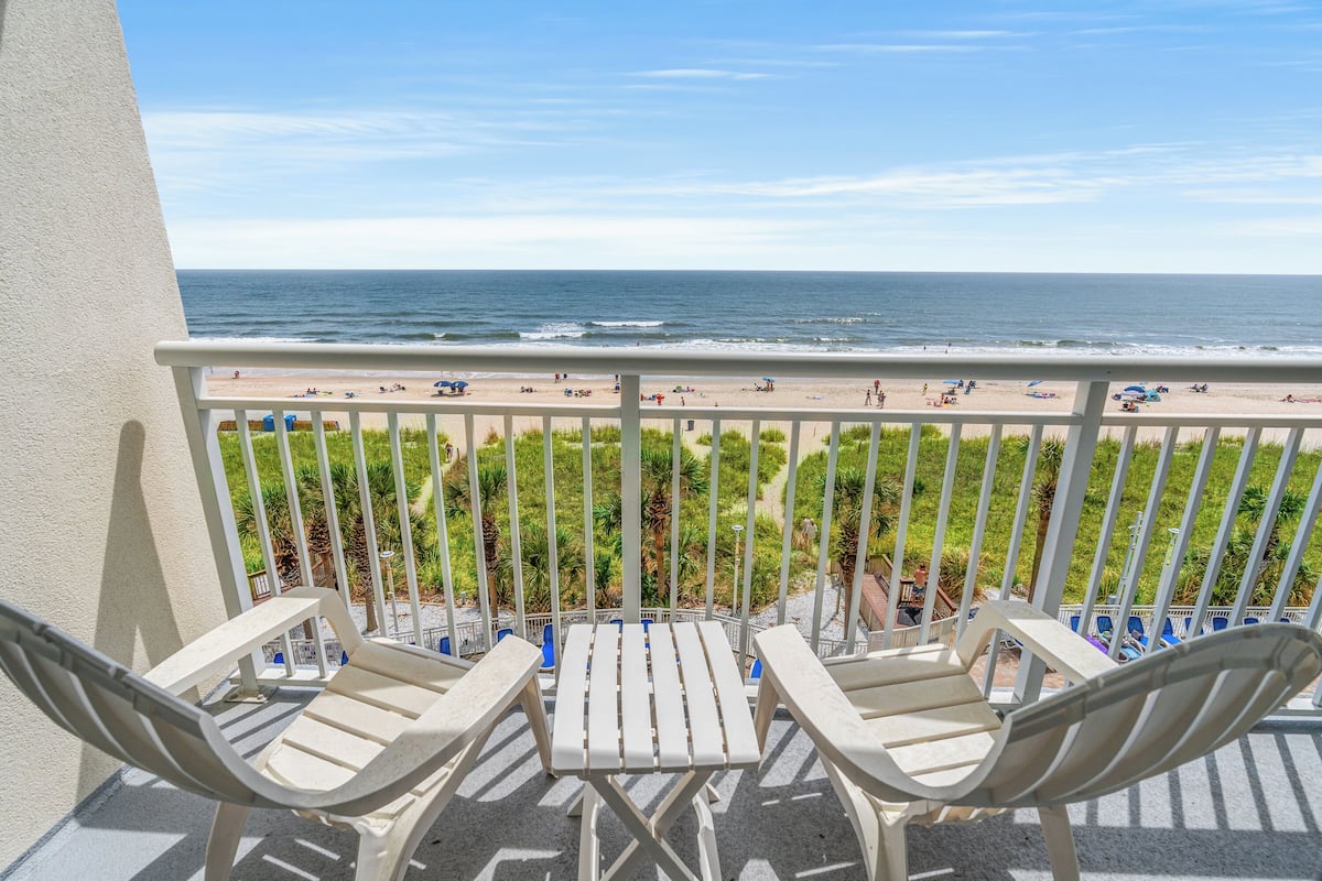 Varanda com duas cadeiras de plástico branco e uma mesa pequena, com vista para uma praia com pessoas e guarda-sóis.