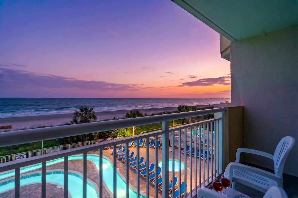 Uma varanda com vista para uma piscina e uma praia ao pôr do sol.