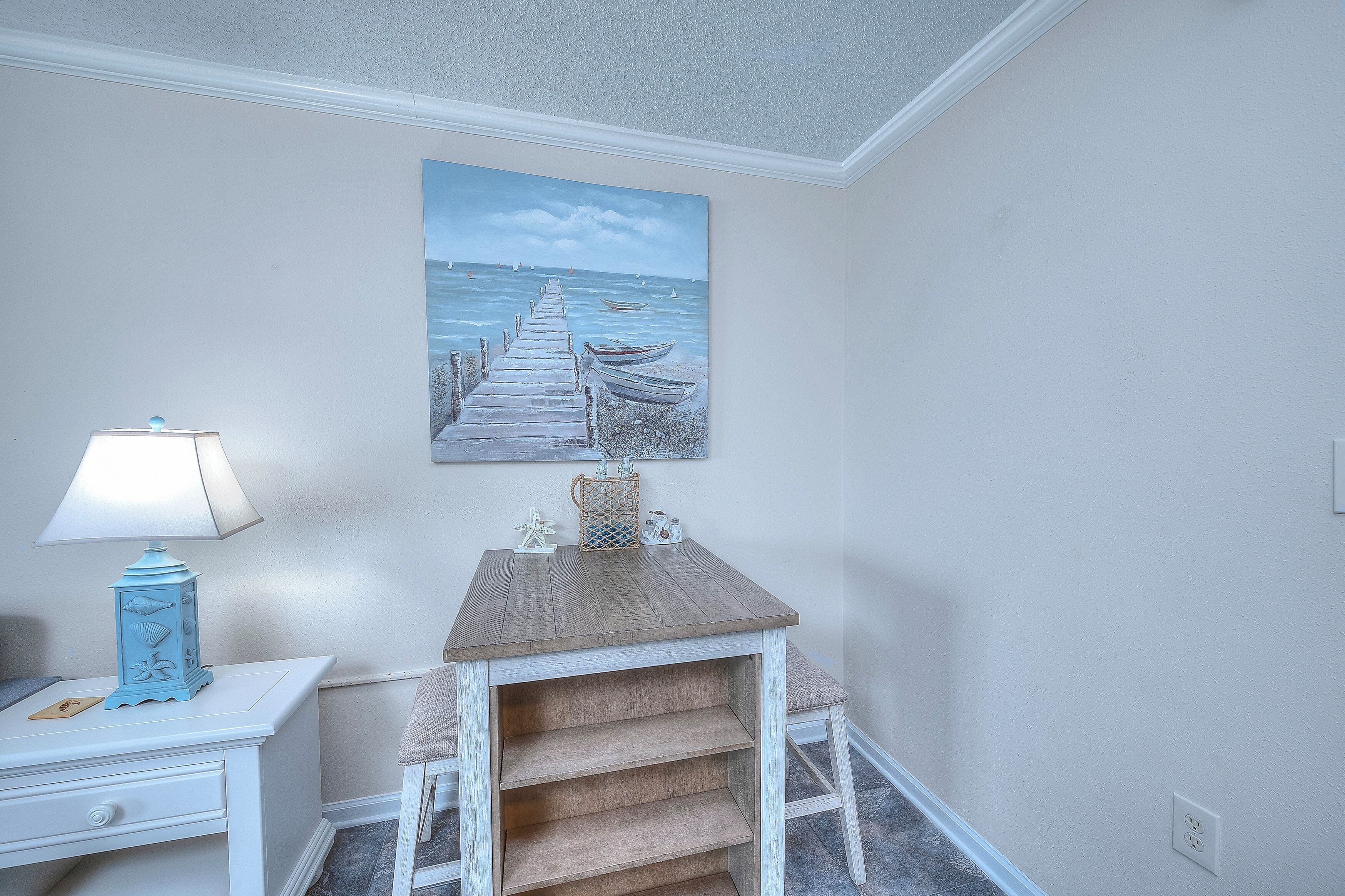 Um quarto com escrivaninha de madeira, luminária, pintura de um cais e uma pequena mesa com luminária.
