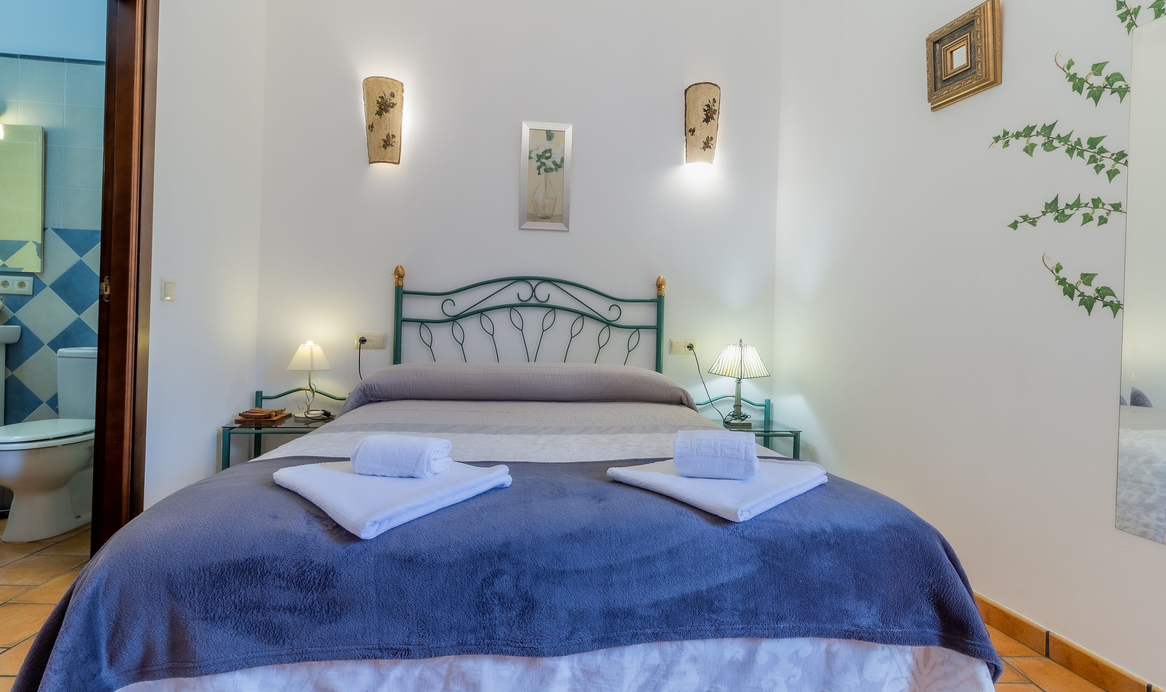 Cama bem arrumada com manta azul, duas mesas de cabeceira com abajures e uma parede com elementos decorativos.