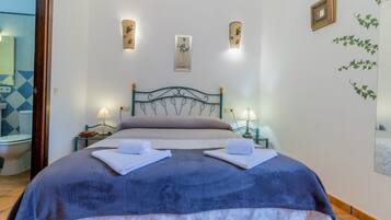 Cama bem arrumada com manta azul, duas mesas de cabeceira com abajures e uma parede com elementos decorativos.