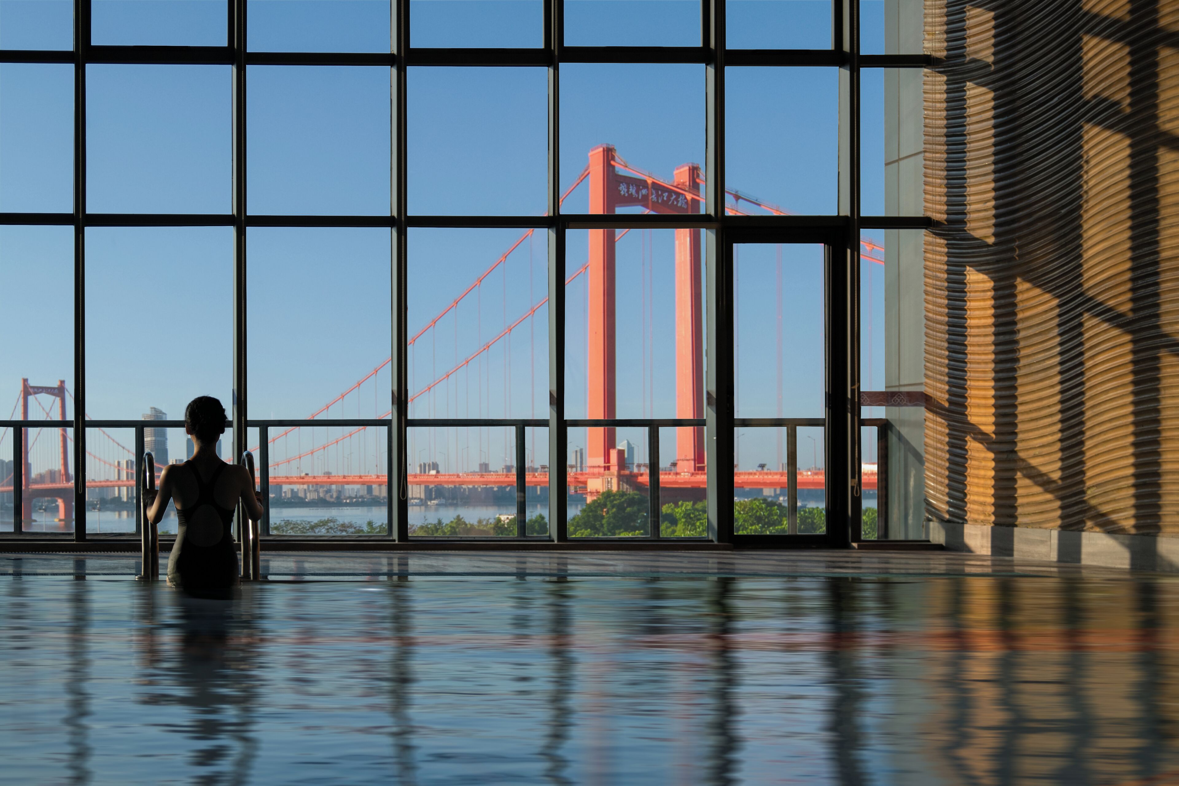 Uma pessoa em uma piscina com vista para uma ponte vermelha e um edifício com grandes janelas.