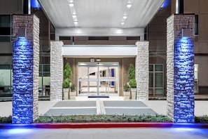 Entrada de hotel com pilares de pedra, portas de vidro e borda de calçada vermelha.