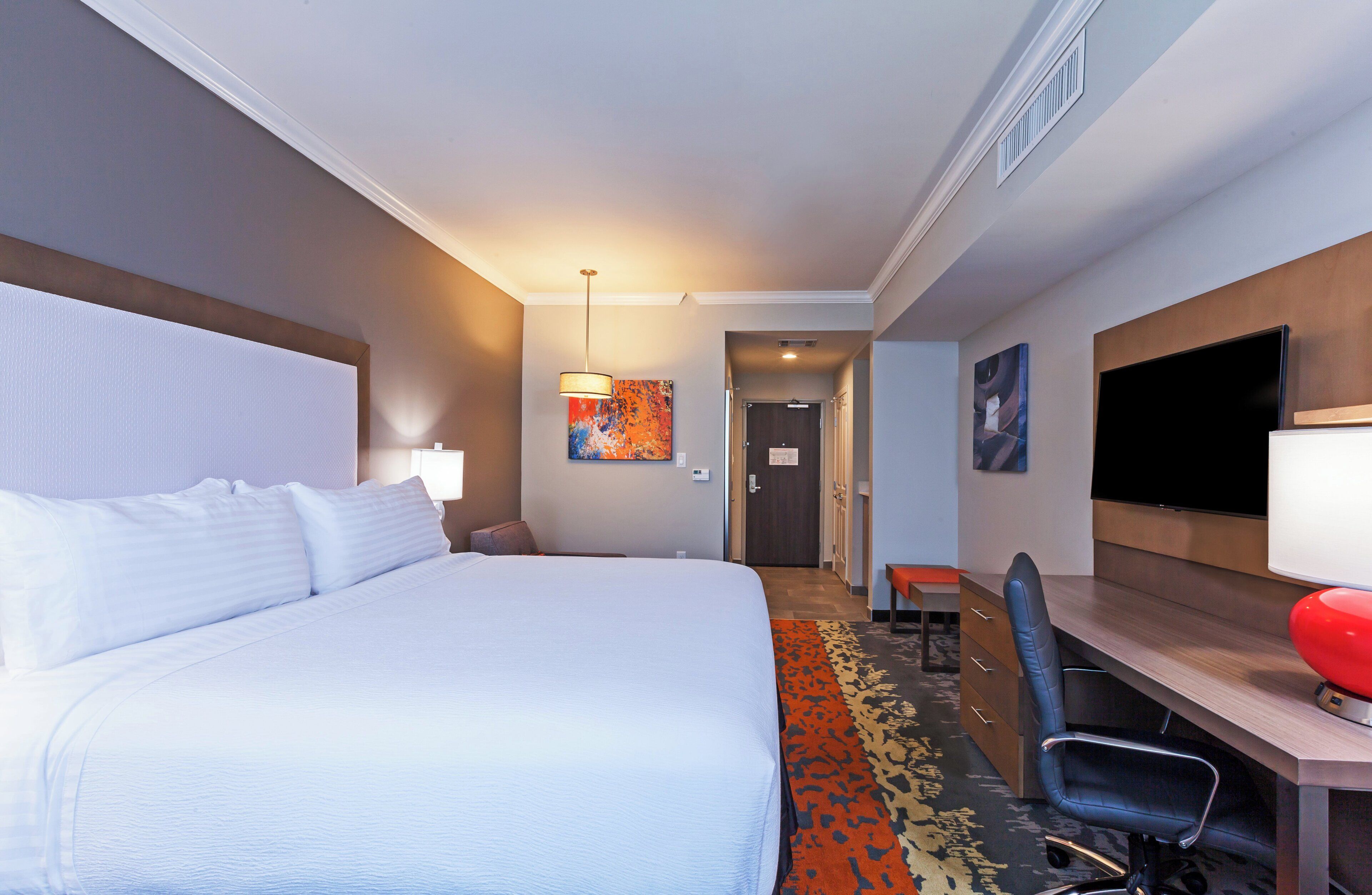 Quarto de hotel com uma cama grande, uma escrivaninha com cadeira, televisão e um quadro na parede.