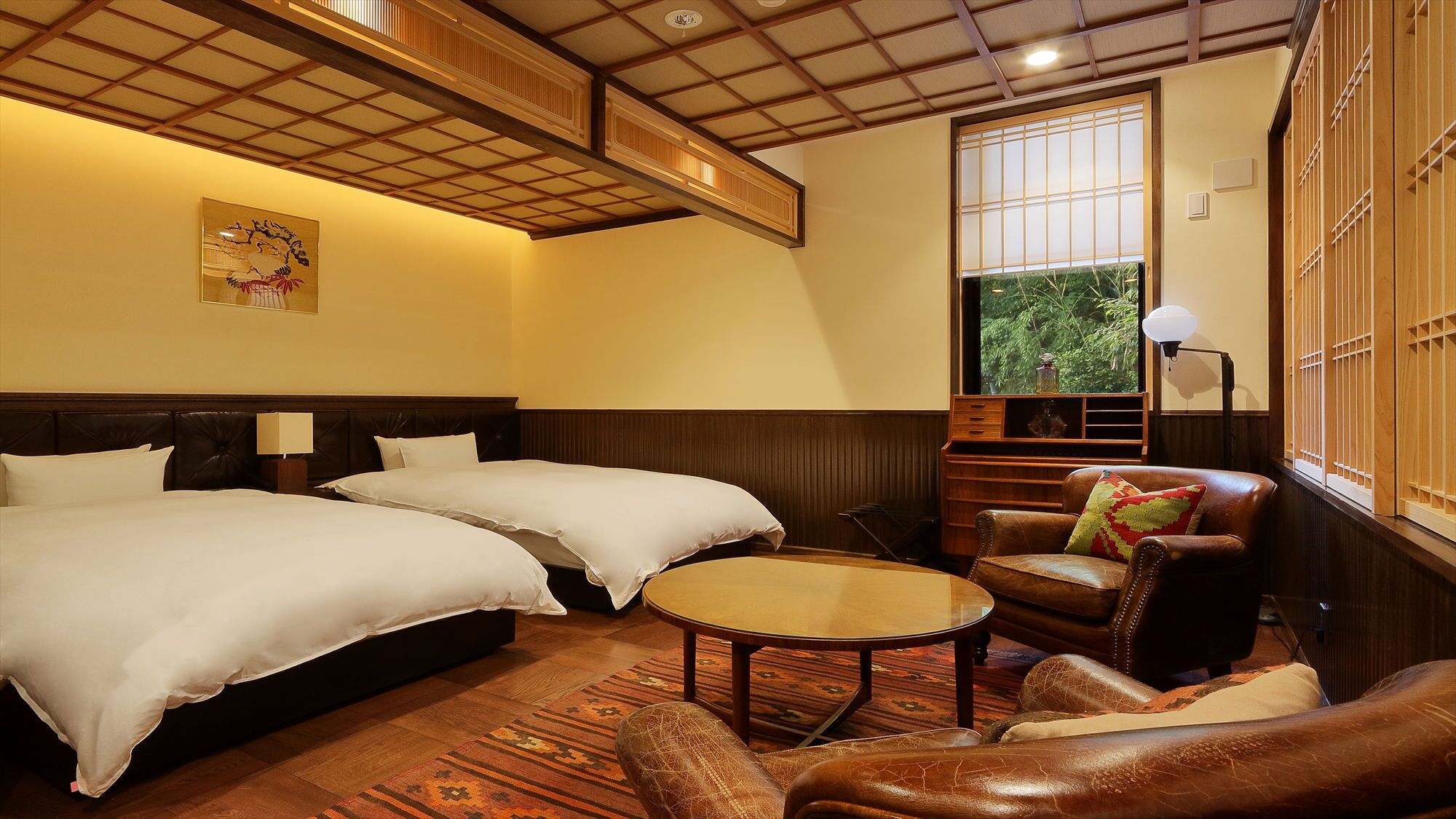 Quarto de hotel com duas camas, uma mesa redonda pequena, uma poltrona de couro, um armário de madeira e uma janela grande com vista para áreas verdes.