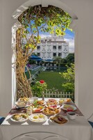 Mesa de café da manhã com diversos pratos e uma rosa, com vista para um jardim e um edifício branco.