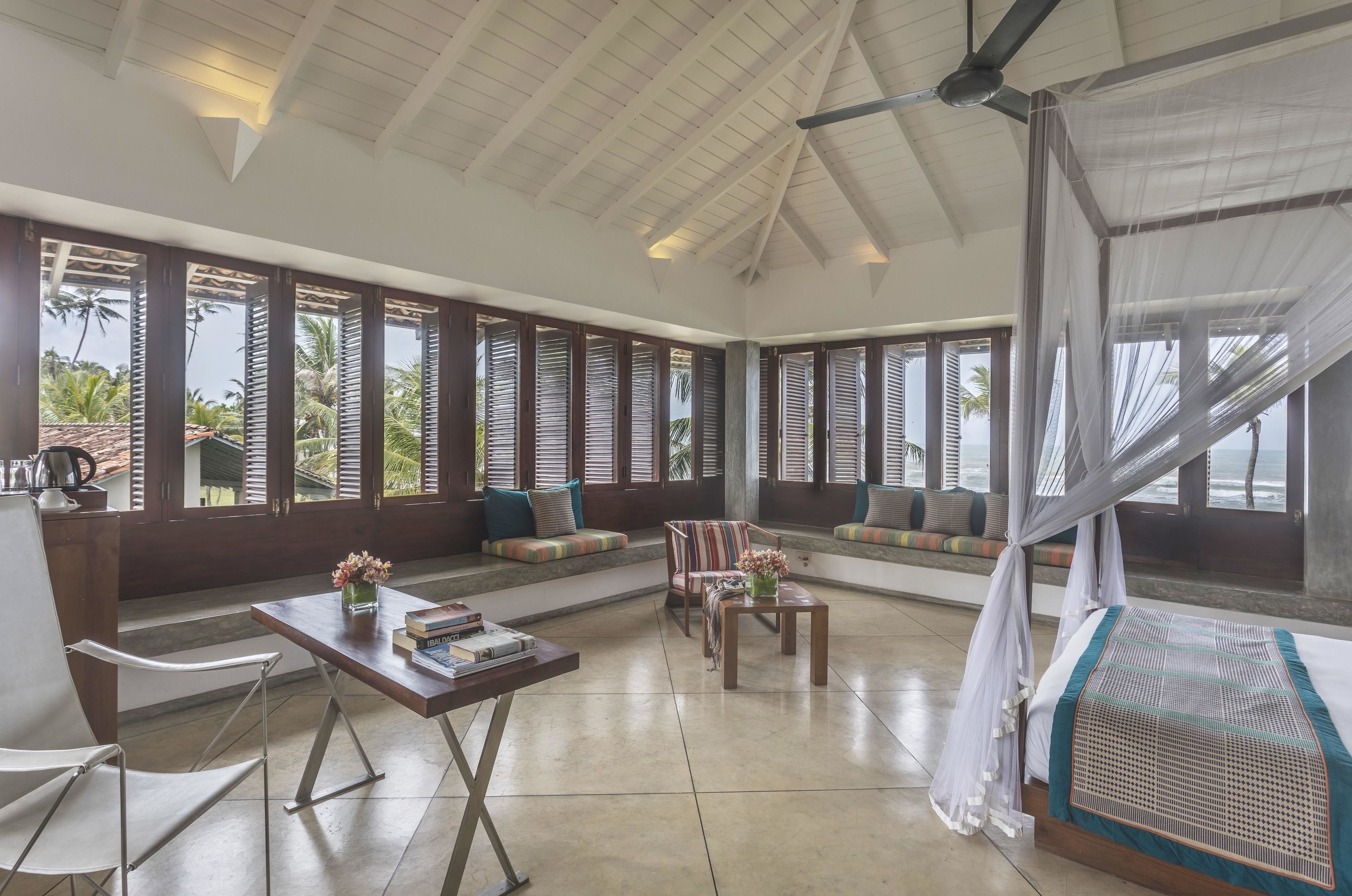 Um quarto espaçoso com teto alto, grandes janelas com venezianas de madeira, uma cama com mosquiteiro, um conjunto de sofás, uma mesa com livros e um vaso de flores, e uma cadeira.