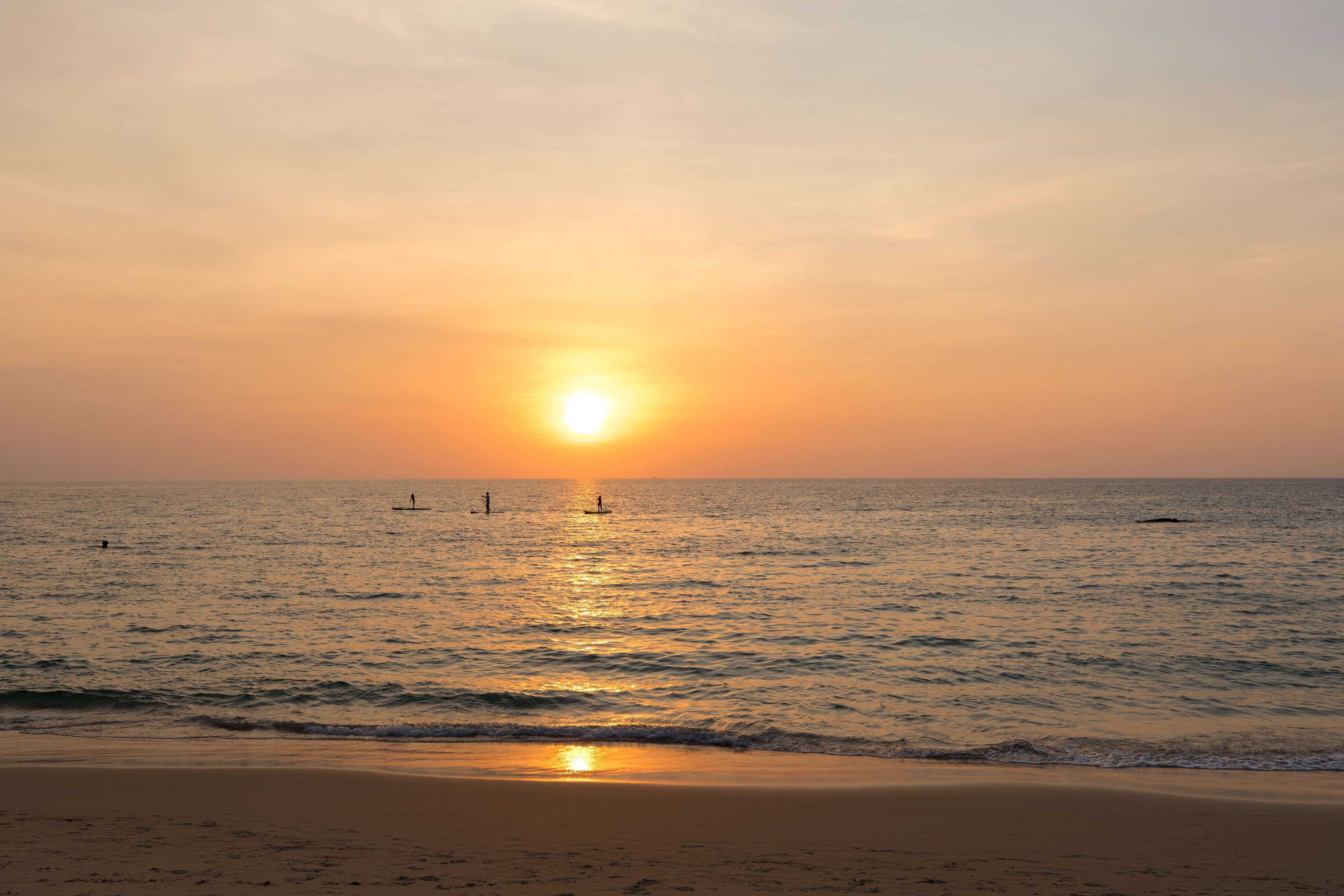 Uma praia com o sol se pondo sobre o oceano, águas calmas e uma orla arenosa.