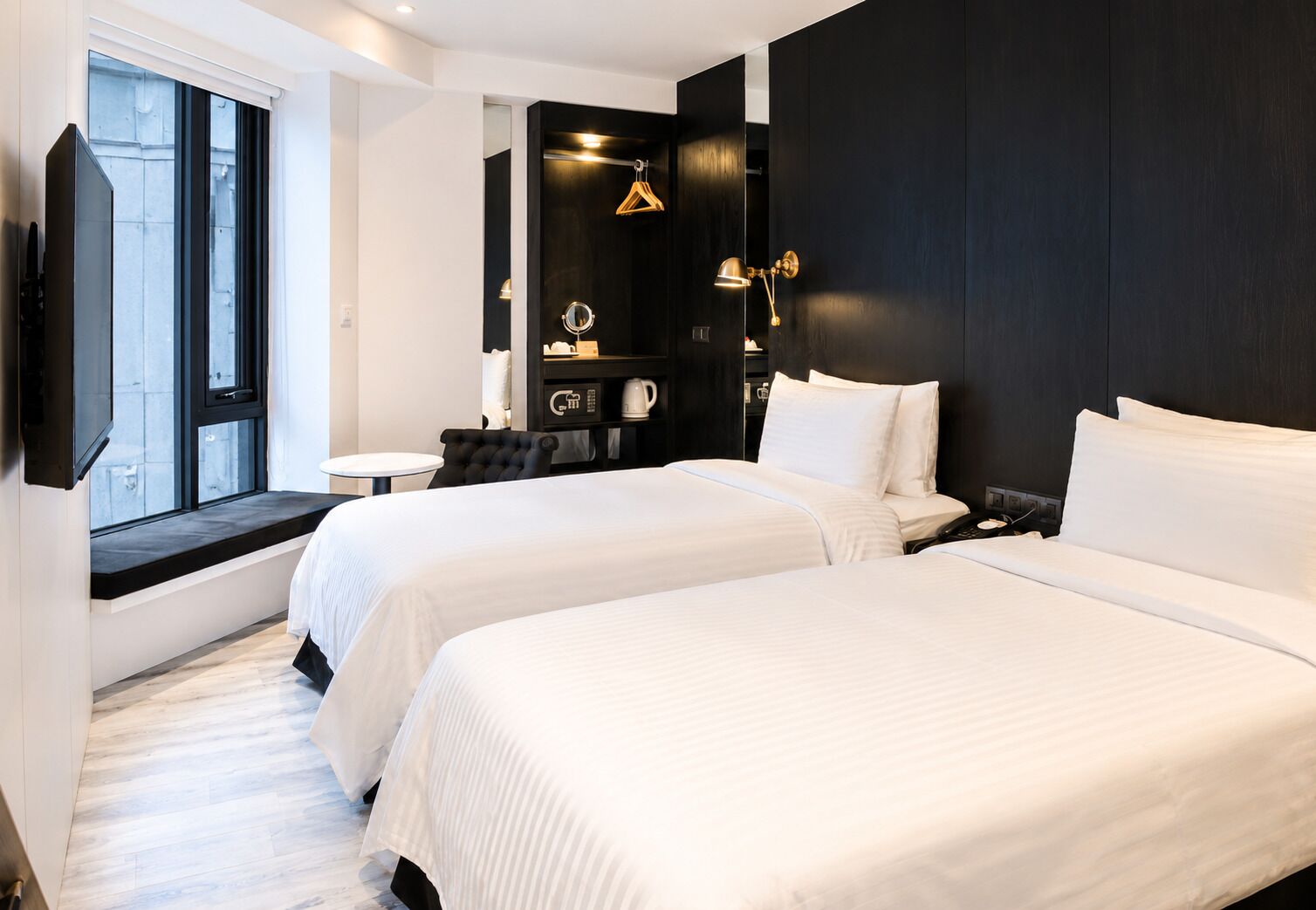 Quarto de hotel com duas camas, uma mesa pequena, uma cadeira, uma janela e uma parede de destaque escura.