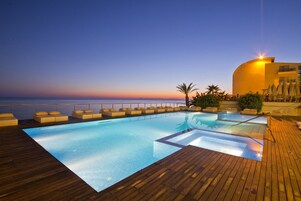 Área de piscina com vista para o oceano ao pôr do sol, deck de madeira e assentos de descanso.