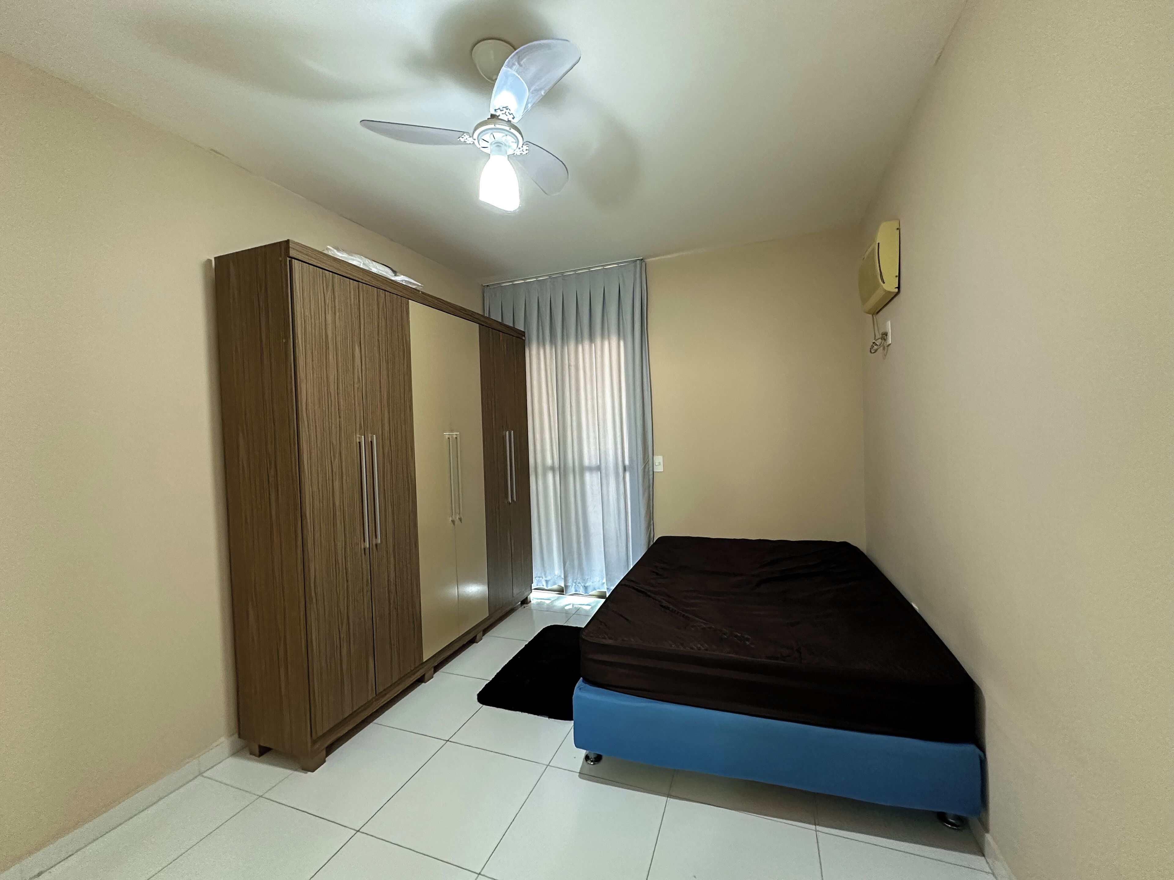 Um quarto pequeno e bem iluminado, com cama, guarda-roupa e ventoinha no teto.