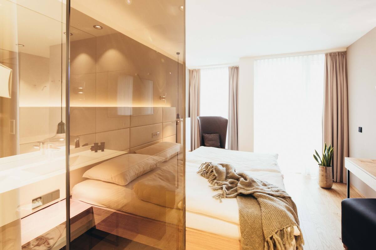 Quarto de hotel moderno com divisória de vidro separando o banheiro do quarto. O banheiro possui um espelho grande, pia e bancada. O quarto tem uma cama com manta, uma cadeira e um vaso de plantas.