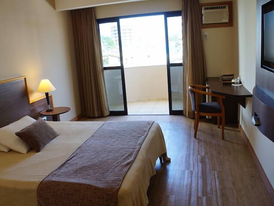 Quarto de hotel com uma cama grande, uma mesa, uma cadeira, uma televisão e uma janela com cortinas.