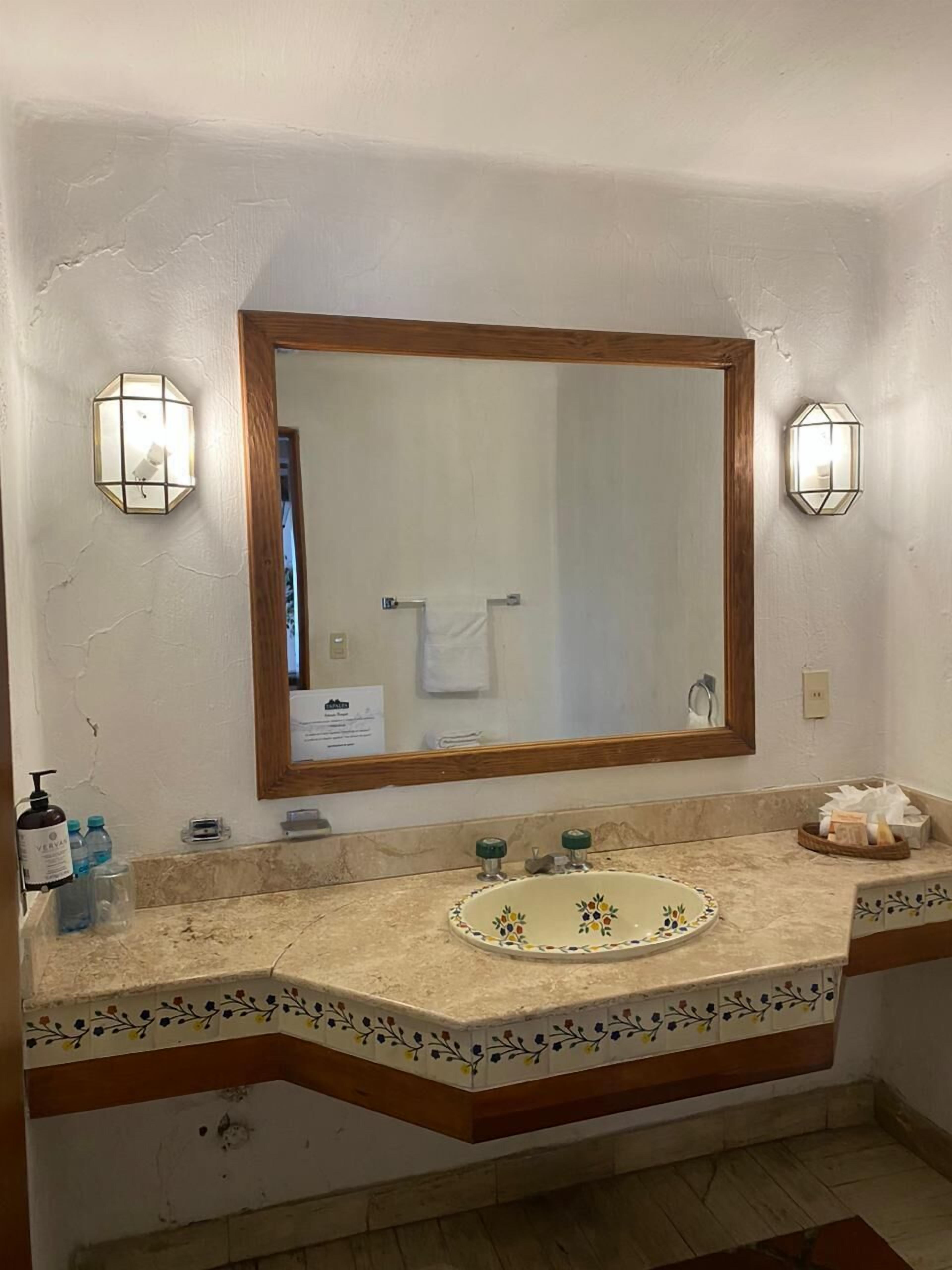 Um banheiro com um espelho grande, duas luminárias na parede, uma pia com cuba decorativa e produtos de higiene no balcão.
