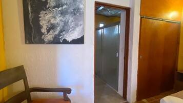 Um quarto com teto de tijolos, uma cama, uma cadeira, um quadro e uma porta que dá para outro espaço.