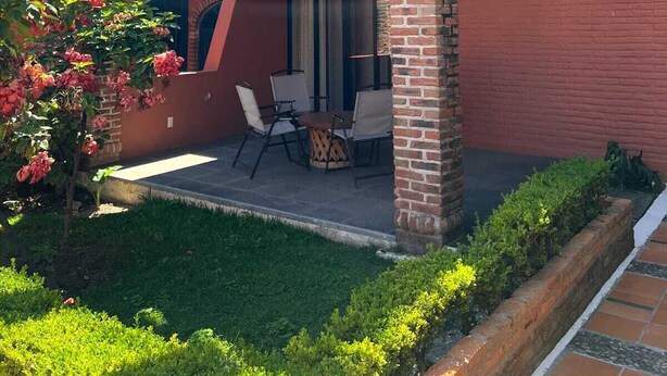 Um edifício de dois andares com varanda, um pátio com cadeiras e um jardim.