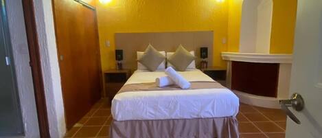 Quarto de hotel com teto de tijolos, cama com lençóis brancos, mesa de cabeceira com abajur e lareira fixada na parede.