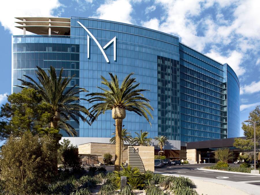 Um hotel moderno com um logotipo 'M' em destaque, rodeado por palmeiras e um jardim bem cuidado.
