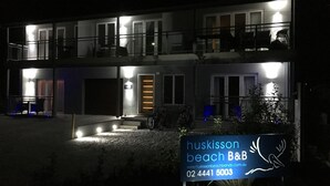 Um edifício de dois andares com janelas e portas iluminadas, uma placa com o nome 'Huskisson Beach B&B' e uma bicicleta estacionada em frente.