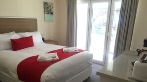 Quarto de hotel com uma cama grande, uma mesa de cabeceira, um quadro na parede e vista pela porta de vidro.