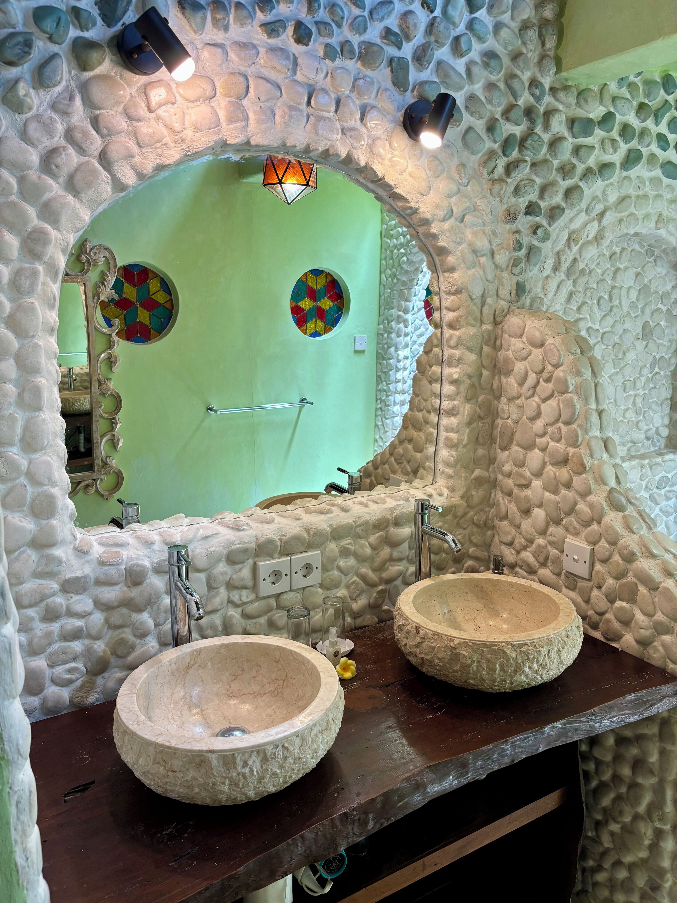 Casa de banho com parede em pedra, duas pias e um espelho com vitrais coloridos.
