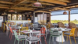 Restaurante com cadeiras e mesas coloridas, vigas de madeira no teto e um bar com garrafas e copos.