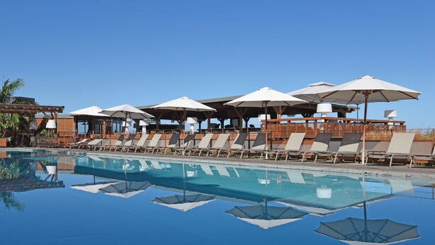Uma área à beira da piscina com espreguiçadeiras e guarda-sóis, uma piscina azul límpida e um deck de madeira.