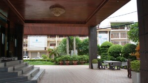 Um espaço coberto ao ar livre com piso de tijolos, degraus que levam à entrada de um edifício e vasos com plantas.