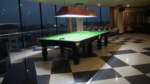 Uma mesa de bilhar numa sala com chão xadrez, grandes janelas e vista para a cidade.