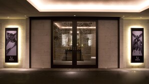 Entrada de hotel com porta de vidro, emoldurada por paredes de pedra, e iluminada por luzes no teto.