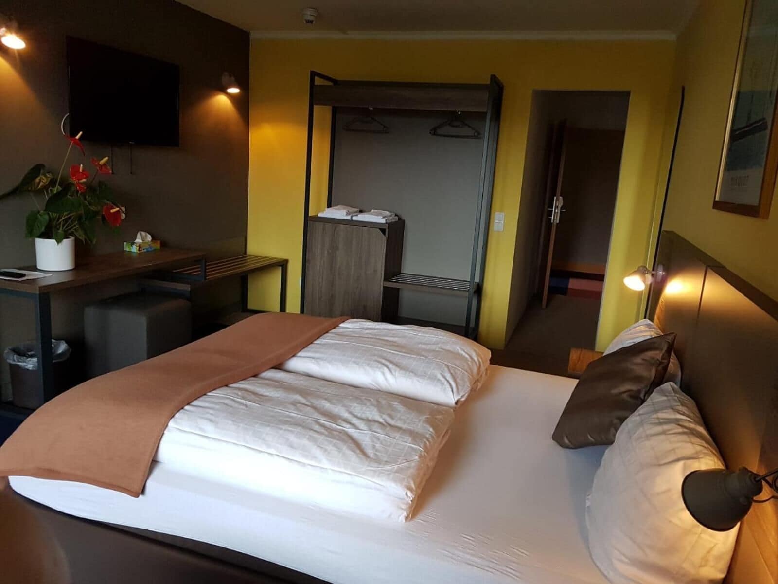 Quarto de hotel com cama, mesa de cabeceira e escrivaninha com cadeira. Há um espelho na parede, uma planta e uma TV fixada na parede.