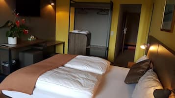 Quarto de hotel com cama, mesa de cabeceira e escrivaninha com cadeira. Há um espelho na parede, uma planta e uma TV fixada na parede.
