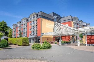 Hotel com vários andares, entrada coberta e área ajardinada com arbustos.