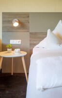 Quarto de hotel com uma cama, uma mesinha redonda com uma chávena e uma planta, e um candeeiro fixado na parede.