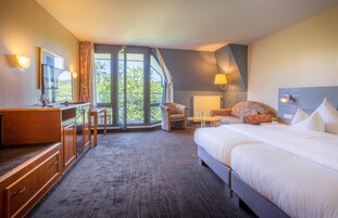 Quarto de hotel com uma cama grande, uma escrivaninha com cadeira, uma mesinha e uma janela com cortinas.