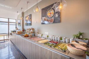 Disposição de buffet com diversos alimentos, utensílios de servir, uma grande janela e luminárias pendentes.