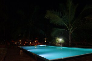 Piscina à noite com palmeiras e um edifício ao fundo.