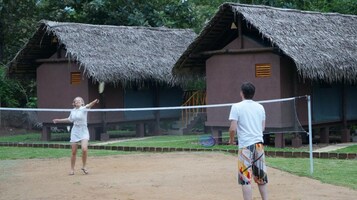 Jogo de voleibol numa área gramada, com ocas em palha ao fundo.