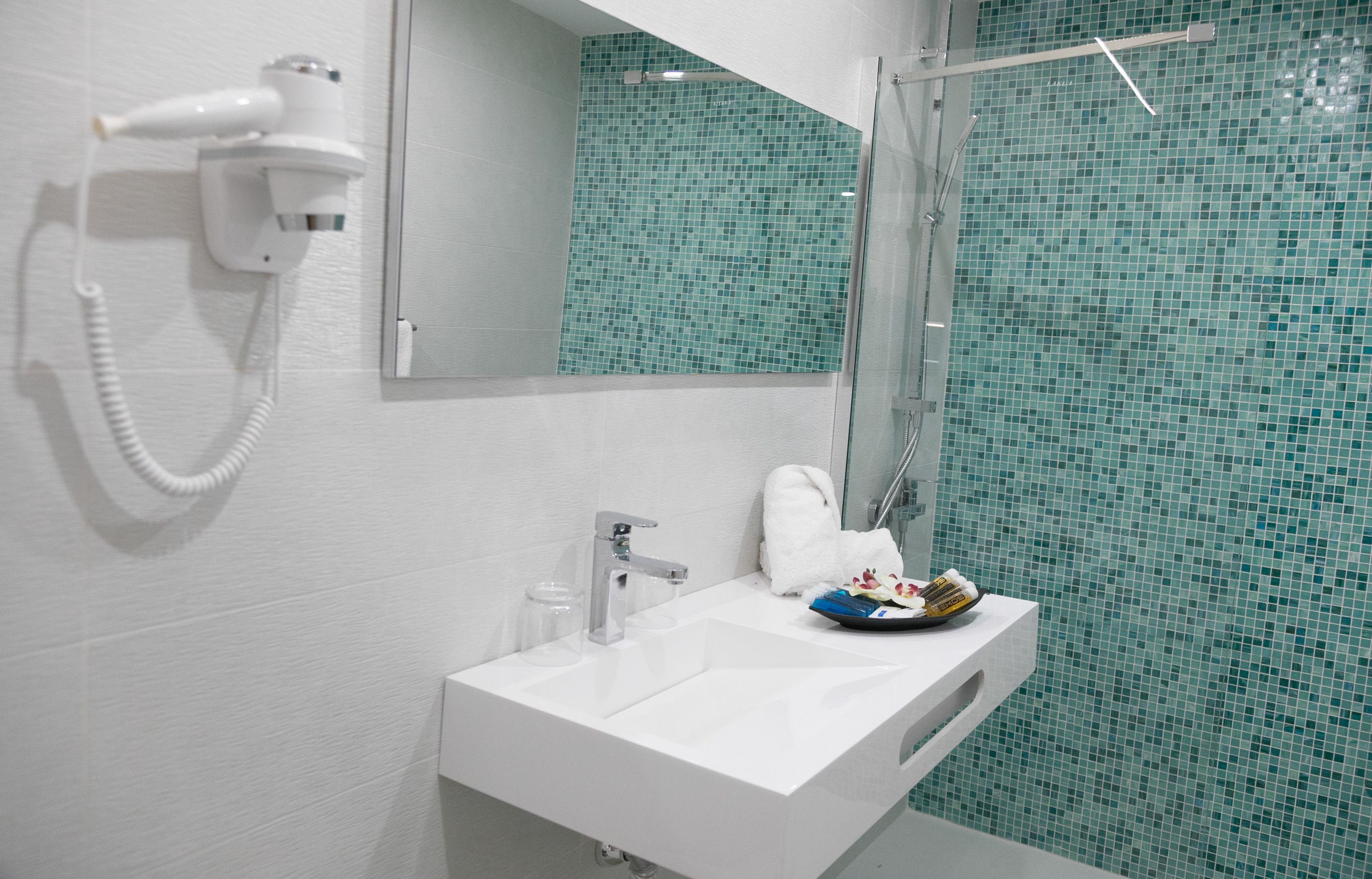 Casa de banho com lavatório branco, secador de cabelo, toalha e cabine de duche em vidro.