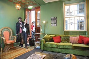 Um quarto aconchegante com um sofá verde, uma cadeira de vime e um letreiro que diz 'É HORA DE TOMAR CHAMPAGNE E DANÇAR NA MESA'.