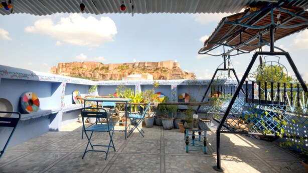 Terraço no telhado com vista para uma fortaleza histórica, cadeiras azuis, plantas em vasos e um balanço.