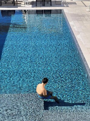 Um homem sentado na beira de uma piscina azul cristalina, com chão de azulejos e móveis de jardim ao fundo.