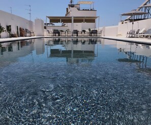 Piscina cristalina com cadeiras de praia e guarda-sóis, cercada por paredes brancas e um edifício com telhado plano.