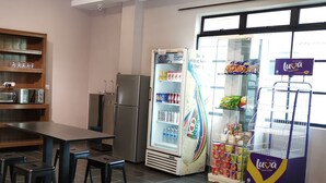 Uma pequena cozinha compacta com geladeira, micro-ondas e torradeira. Há uma máquina de vendas automática e uma vitrine com lanches.