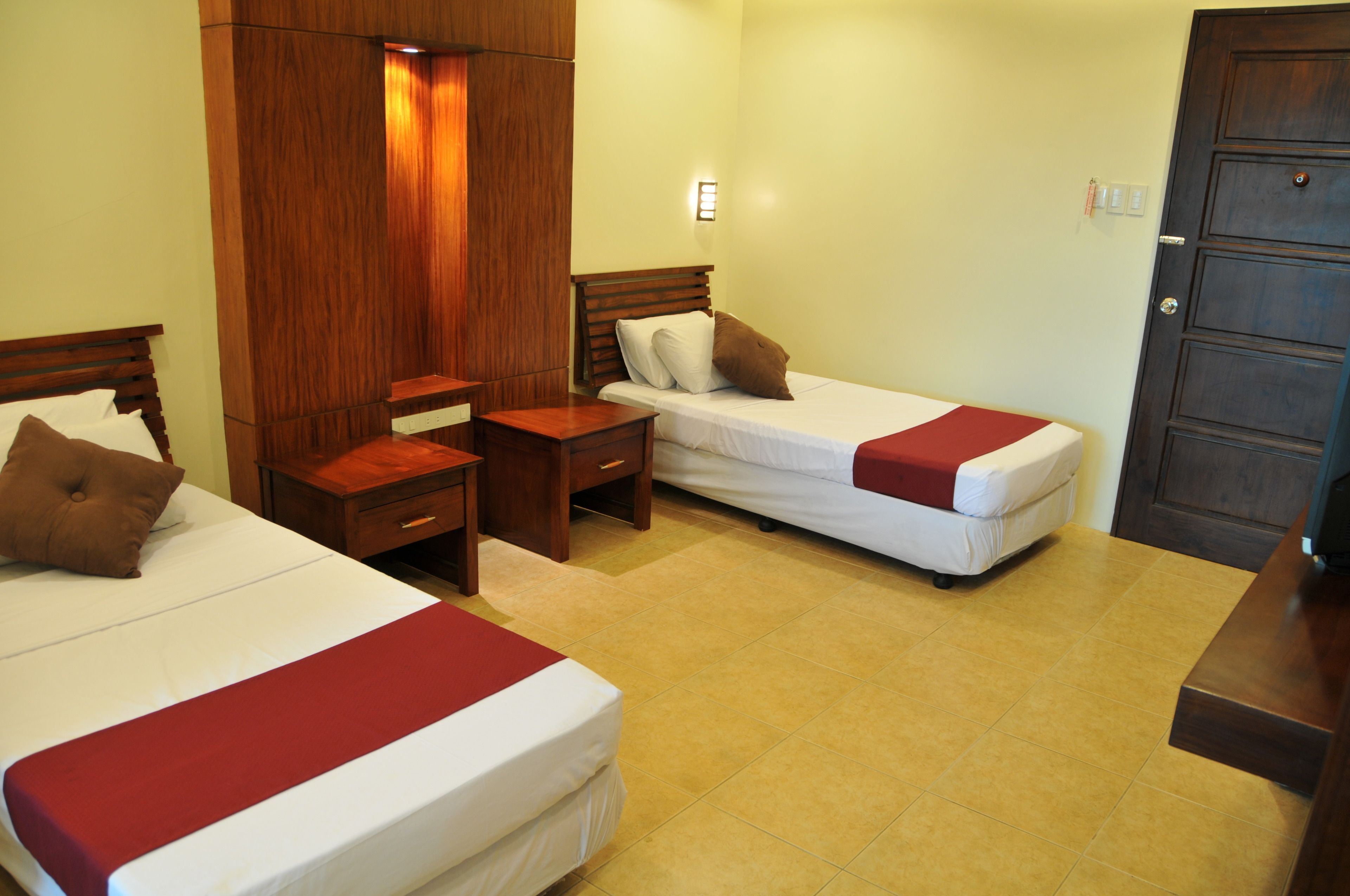 Quarto de hotel com duas camas, cabeceiras de madeira, uma mesinha de cabeceira, uma pequena mesa e um guarda-roupa.