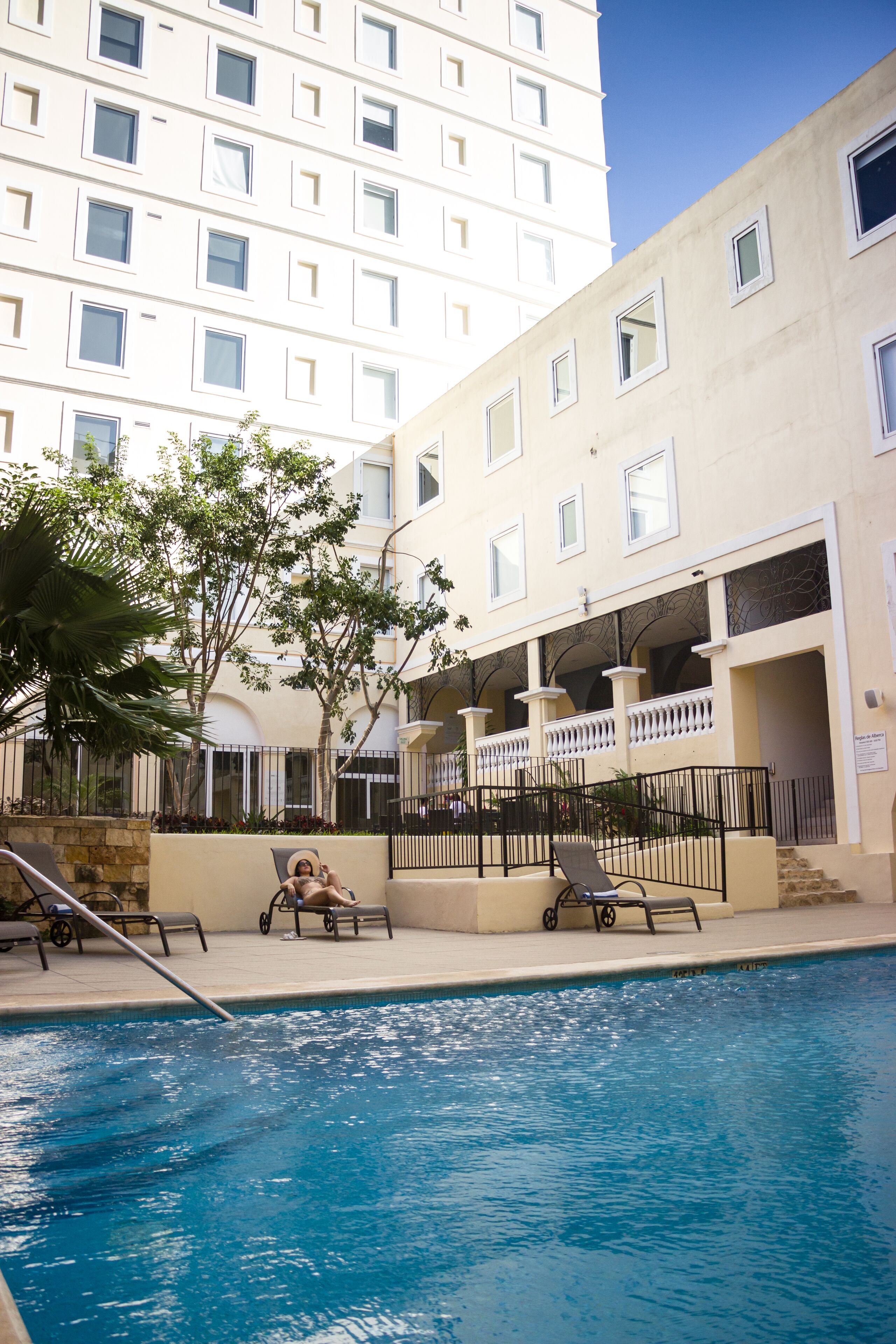Piscina com espreguiçadeiras e um edifício com varandas ao fundo.