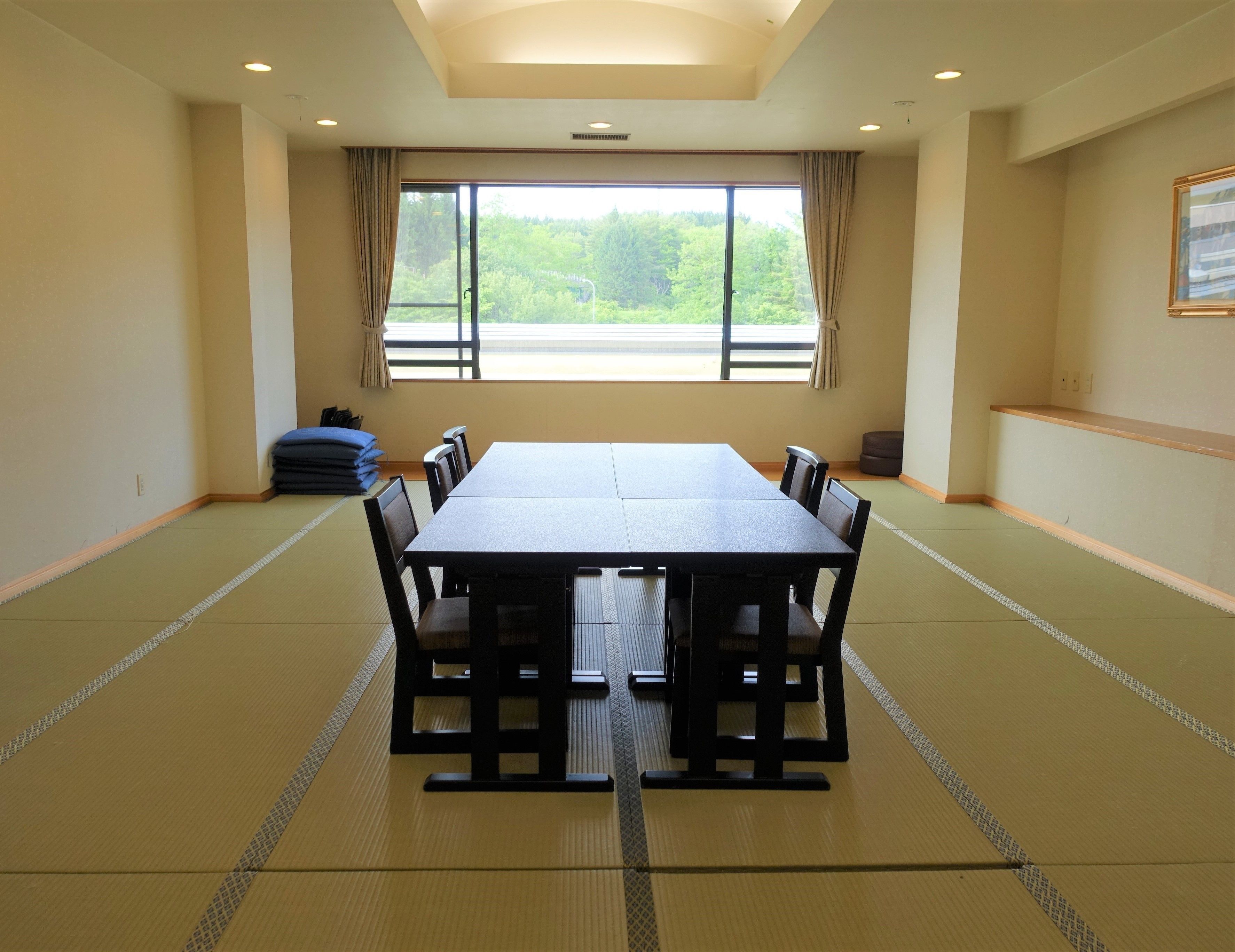 Uma sala de conferências com uma mesa comprida, cadeiras, tatames, uma janela grande com cortinas e um quadro na parede.
