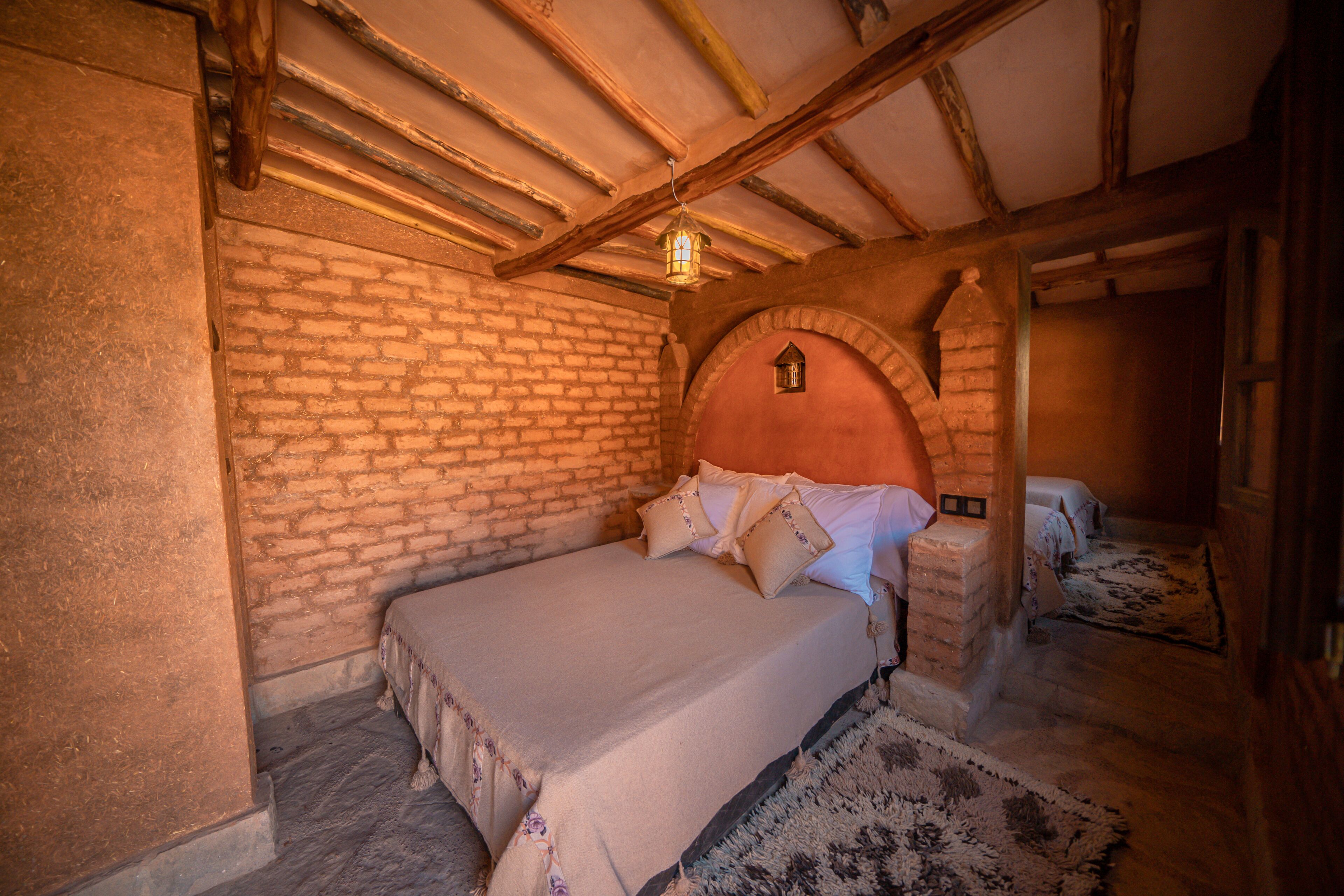 Quarto com parede de tijolos, teto de madeira, cama com colcha branca e uma pequena janela emoldurada por madeira.