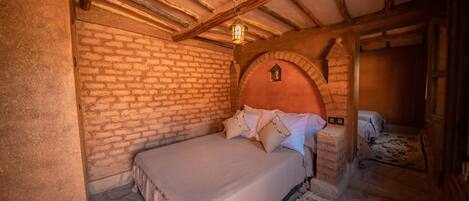 Quarto com parede de tijolos, teto de madeira, cama com colcha branca e uma pequena janela emoldurada por madeira.